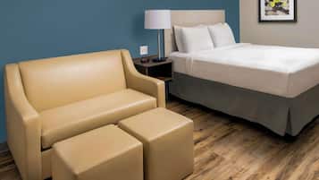 Quarto de hotel com um sofá bege, um banquinho a combinar, uma cama com roupa de cama branca, uma mesinha de cabeceira com abajur e um quadro pendurado na parede.