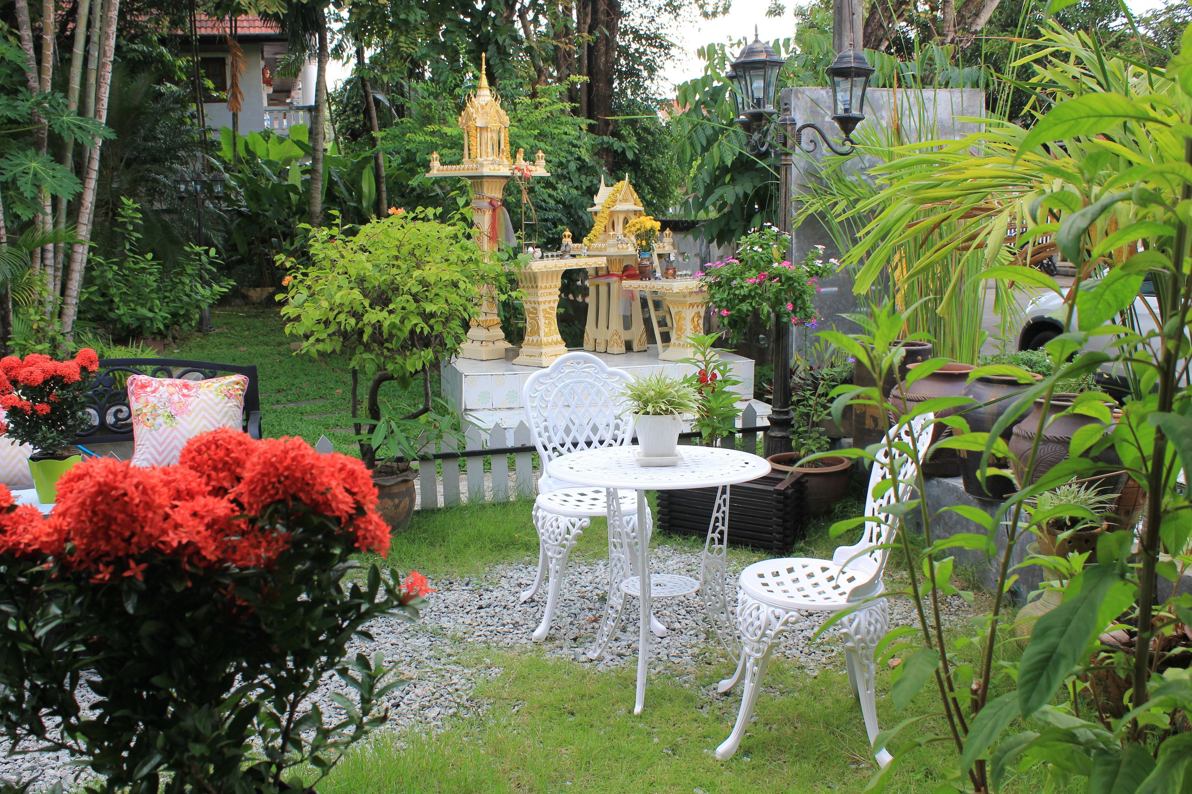 Um jardim com uma mesa e cadeiras de metal branco, rodeado por plantas em vasos e uma fonte decorativa.