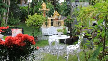 Um jardim com uma mesa e cadeiras de metal branco, rodeado por plantas em vasos e uma fonte decorativa.
