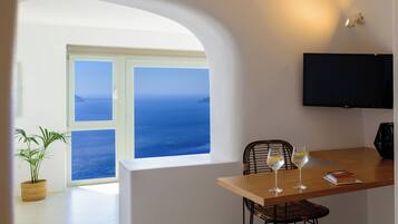Quarto com televisão na parede, mesa de jantar com duas cadeiras e vista para o mar através de uma grande janela em arco.
