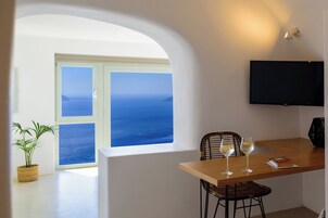 Quarto com televisão na parede, mesa de jantar com duas cadeiras e vista para o mar através de uma grande janela em arco.