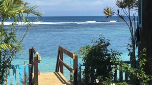 Um deck de madeira leva a uma praia com vista para o oceano, cercado por plantas tropicais e uma construção com telhado de palha.