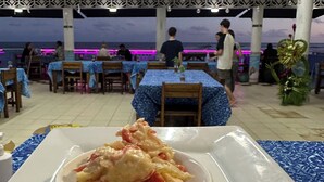 Restaurante com mesas cobertas por toalhas azuis, uma pessoa segurando um prato de comida e uma vista para o mar ao fundo.