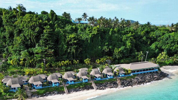 Resort com bangalôs de telhado de palha ao longo da praia, águas turquesa cristalinas e vegetação exuberante ao fundo.