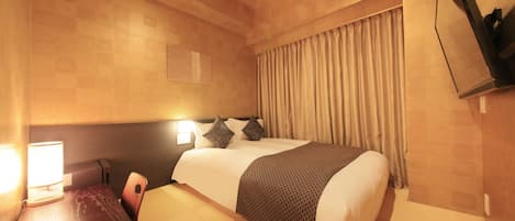 Quarto de hotel com uma cama grande, uma escrivaninha com luminária, uma cadeira e uma televisão na parede.