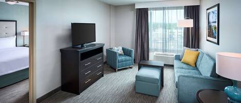 Quarto de hotel com cama, televisão, poltrona azul, sofá, mesa de centro e janela com cortinas.
