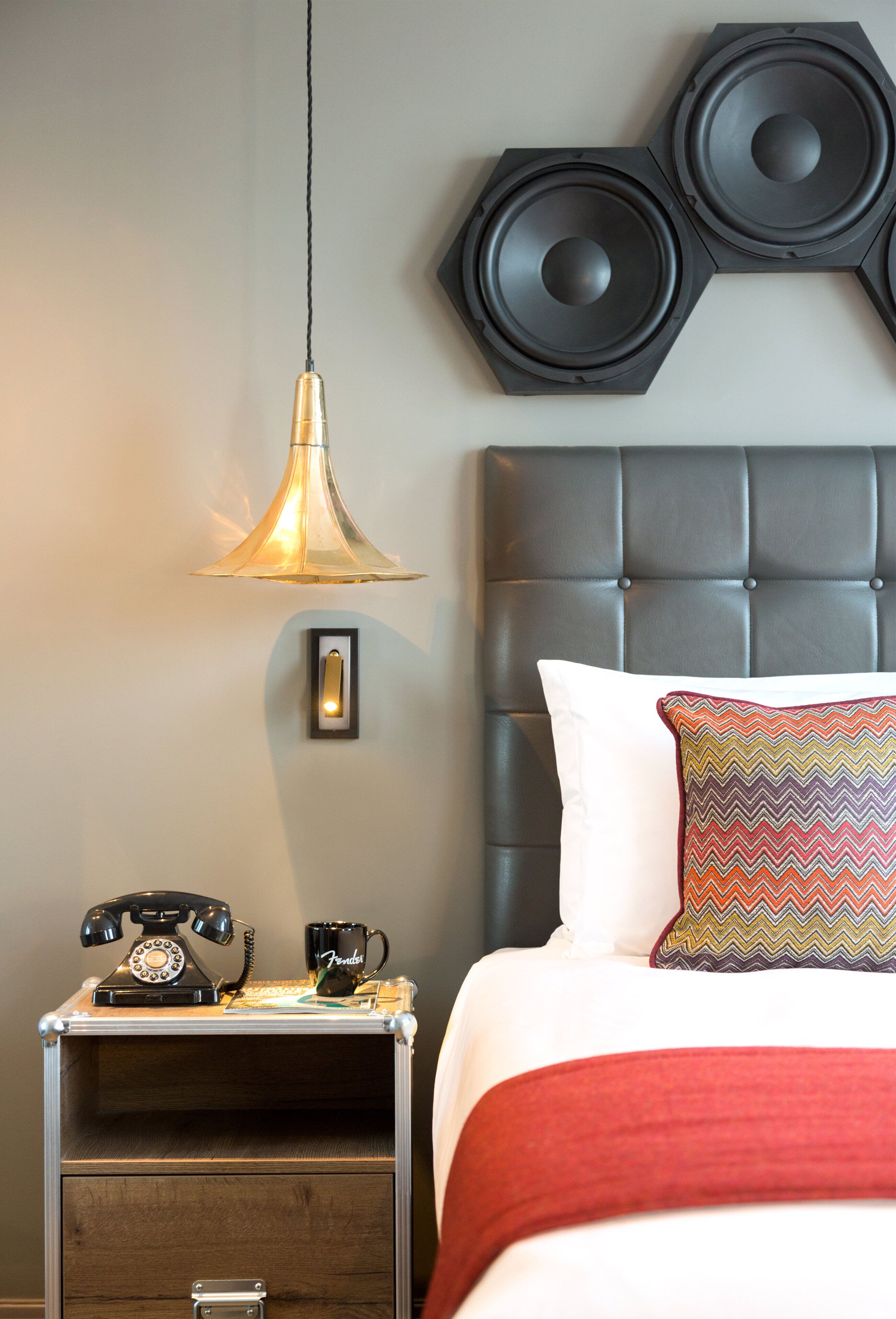Quarto de hotel com cama, mesa de cabeceira com telefone, luminária na parede e painel decorativo.