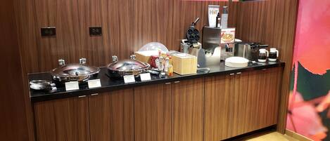 Um balcão de buffet com diversos alimentos e acompanhamentos, incluindo uma máquina de waffles e aparelhos de café.