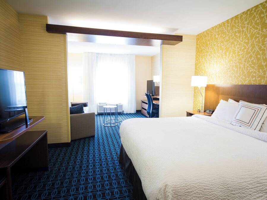 Quarto de hotel com uma cama grande, uma TV em suporte, uma mesa com cadeira e vista para o corredor.