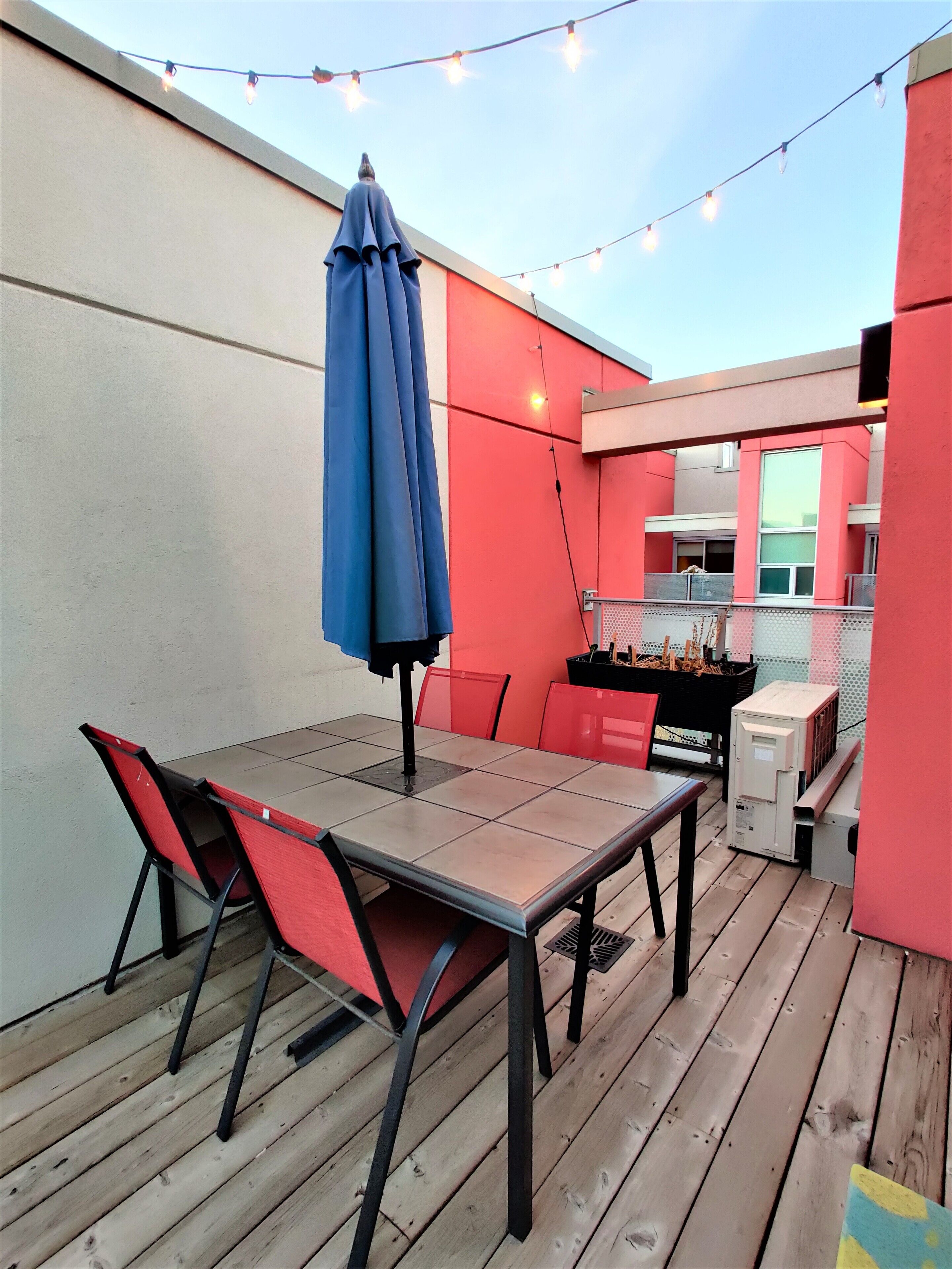 Um pátio externo com mesa de madeira e cadeiras vermelhas, luzes de cordão e um guarda-sol azul.