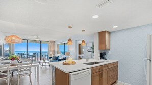 Cozinha moderna com lava-louças, armários de madeira e uma ilha grande. Há uma área de refeições com cadeiras brancas e uma mesa, e vista para o oceano através de grandes janelas.
