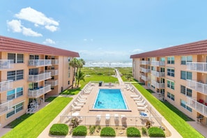 Hotel com piscina cercada por espreguiçadeiras, vista para o oceano e gramado bem cuidado.