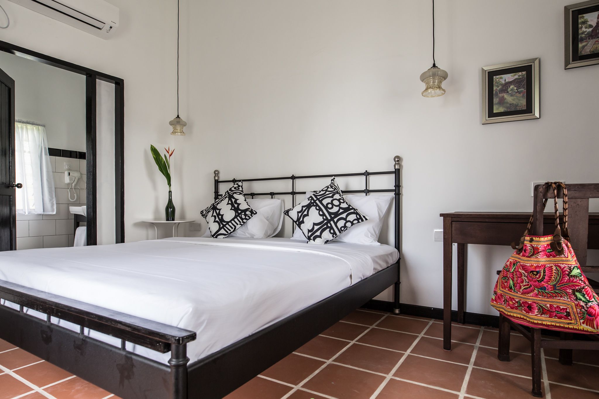 Cama bem arrumada com lençóis brancos e almofadas decorativas, uma pequena mesa de cabeceira com uma planta, uma cadeira de madeira e um quadro na parede.