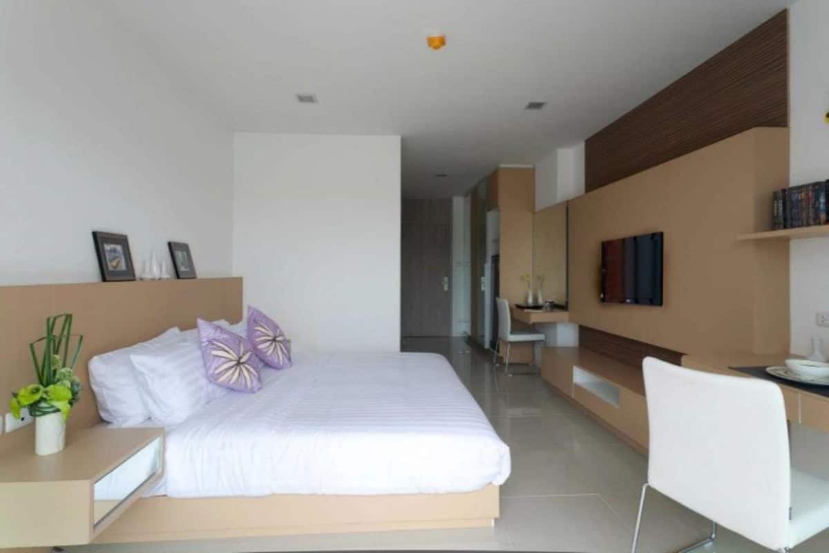 Quarto de hotel moderno com uma cama grande, uma escrivaninha com cadeira, uma televisão na parede e uma pequena planta no criado-mudo.
