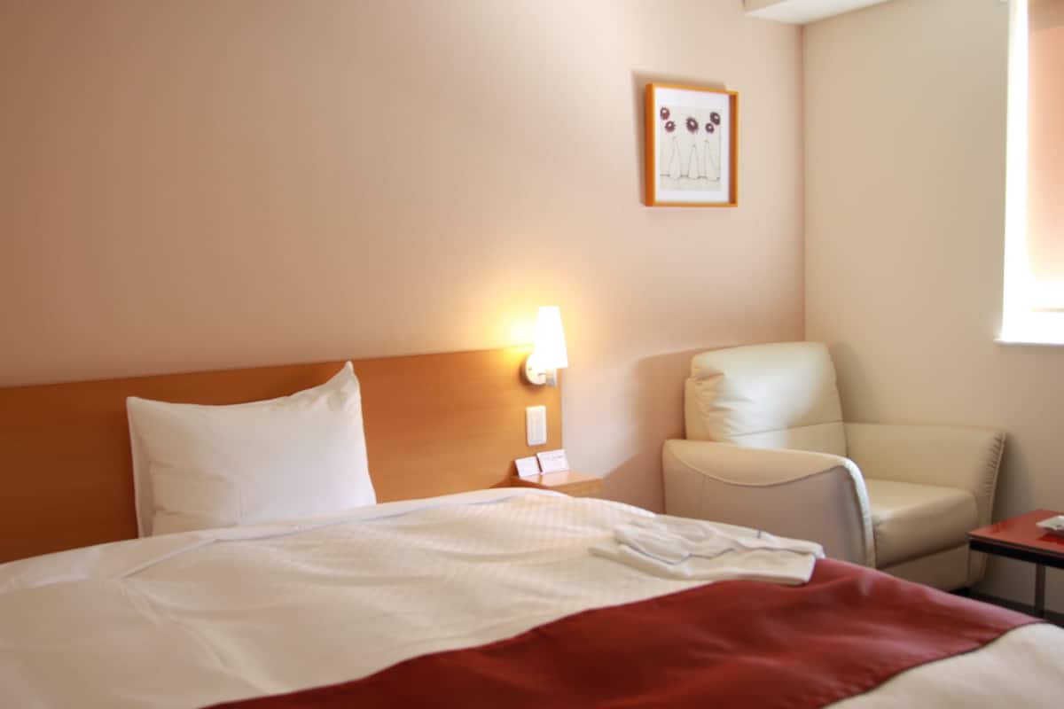 Quarto de hotel com cama, mesa de cabeceira, cadeira e luminária fixada na parede.
