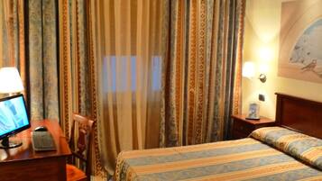Quarto de hotel com cama, escrivaninha com computador, cadeira e mesa de cabeceira com abajur.