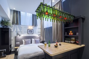 Quarto de hotel moderno com cama, mesa de cabeceira e área de bar com garrafas e copos.