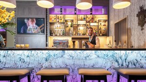 Uma área de bar com uma tela de TV, prateleiras com garrafas de bebidas, um balcão e três banquetas.