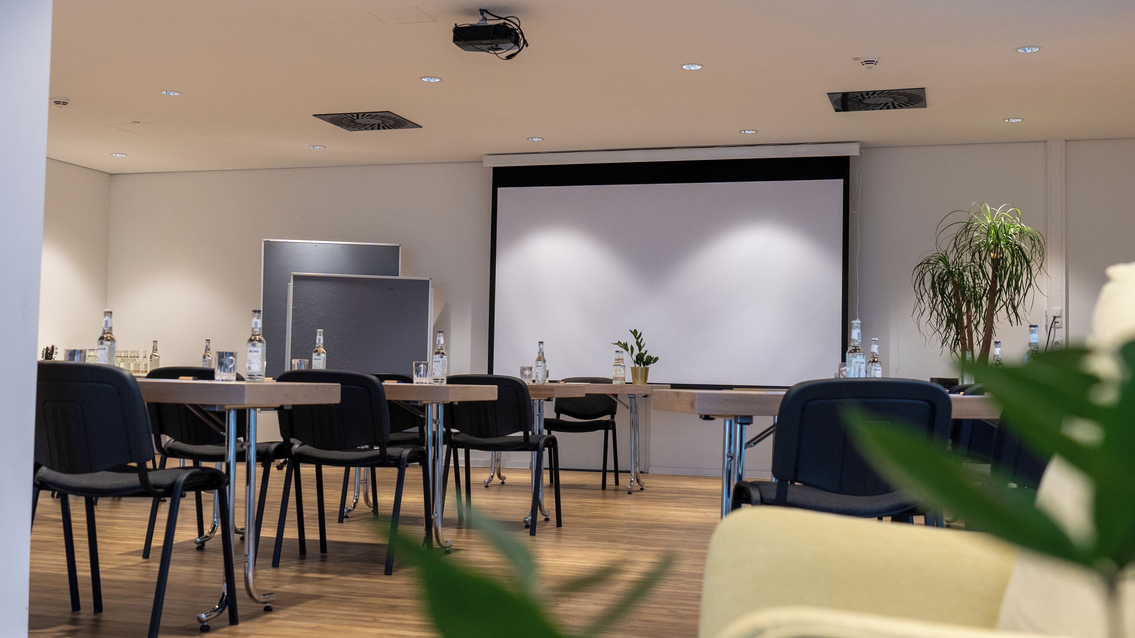Sala de conferências com tela de projetor, mesas, cadeiras e um vaso de planta.