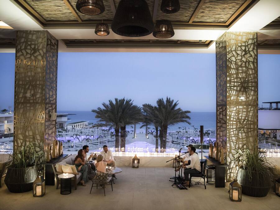 Uma área de estar ao ar livre com vista para a praia e palmeiras, com mesas e cadeiras, e um palco com instrumentos musicais.