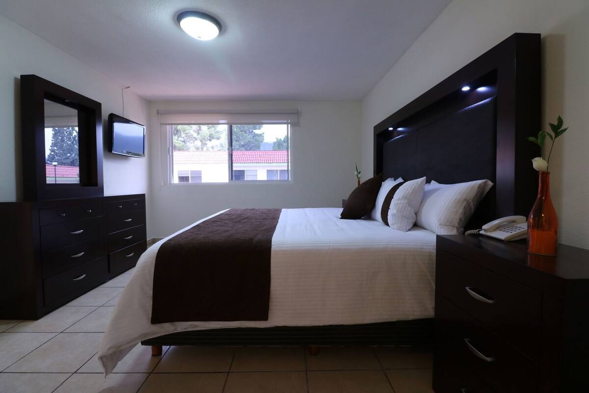 Quarto com cama, penteadeira, espelho, televisão, janela e telefone.