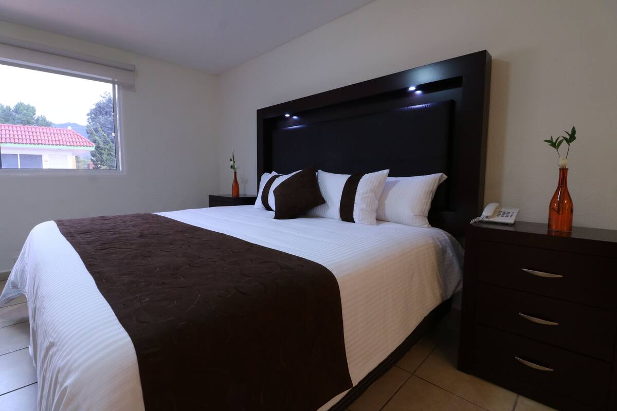 Cama bem arrumada com cabeceira escura, mesa de cabeceira com telefone e janela com vista para um edifício.