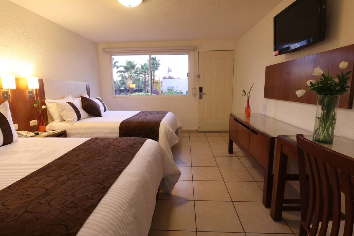 Quarto de hotel com duas camas, televisão, secretária e janela com vista para palmeiras.