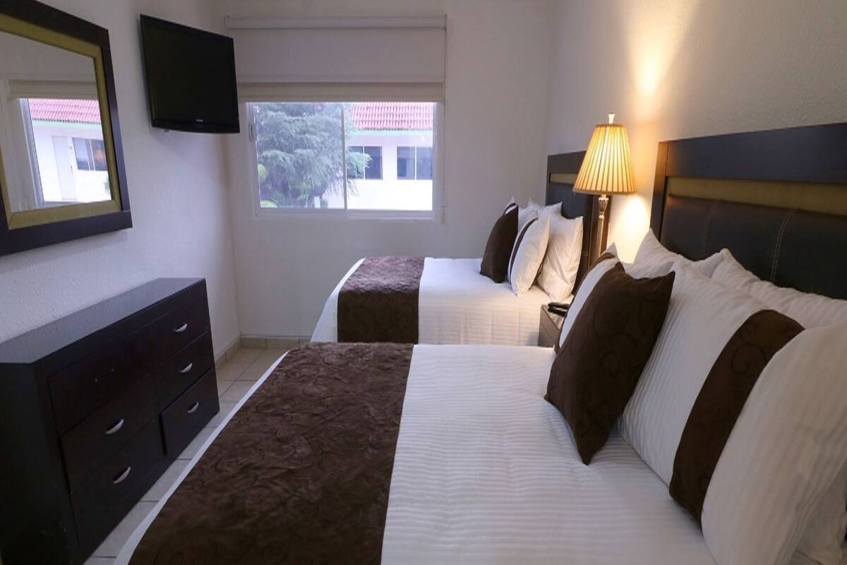 Quarto de hotel com duas camas, uma cômoda, uma televisão e uma janela com vista para árvores e um edifício.