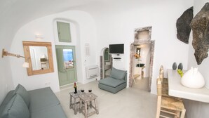 Uma sala de estar moderna com um sofá cinzento, uma mesa de madeira pequena com copos de vinho, uma poltrona cinzenta e uma parede de pedra com uma TV suspensa.