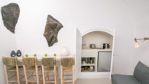Um espaço de estar compacto com área de jantar, apresentando quatro cadeiras de madeira com assentos trançados e uma mesa branca. Uma cozinha compacta é visível, com cafeteira, torradeira e minigeladeira. Um sofá cinza está posicionado no canto.