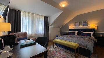 Quarto de hotel com cama, escrivaninha, cadeira, mesa com xícaras, um banco e um tapete colorido.