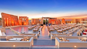 Espaço de refeições no terraço com mesas e cadeiras, piscina e vista para ruínas antigas ao pôr do sol.