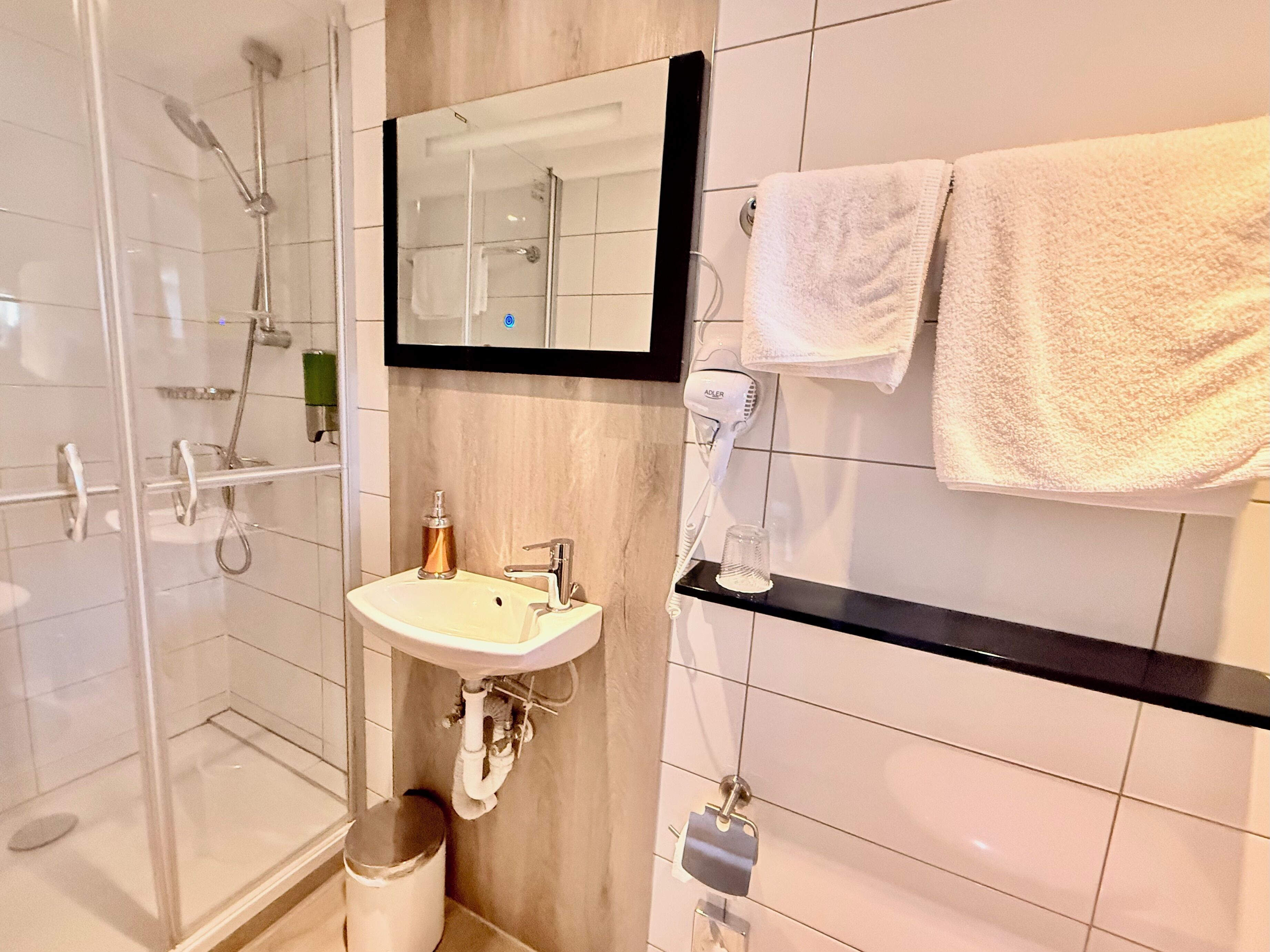 Casa de banho com cabine de duche em vidro, lavatório com dispensador de sabonete, toalheiro com toalhas e secador de cabelo numa prateleira.