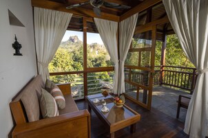 Quarto com sofá de madeira, uma mesa com uma cesta de frutas e vista para uma paisagem montanhosa através de grandes janelas.