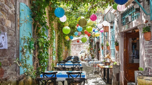Rua estreita alinhada com mesas e cadeiras, coberta por toalhas de mesa azuis, e decorada com lanternas de papel penduradas.