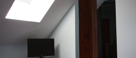Um quarto com um claraboia, uma televisão sobre um móvel de madeira e uma porta que dá para outro cômodo.