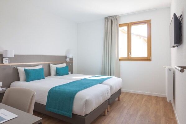 Quarto de hotel com uma cama grande, uma escrivaninha, uma cadeira, uma televisão na parede e uma janela com cortinas.