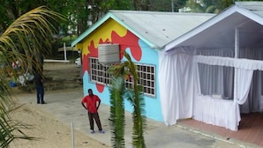 Uma área arenosa ao ar livre com um poço de fogueira de pedra, um pequeno edifício com um mural colorido e uma tenda.