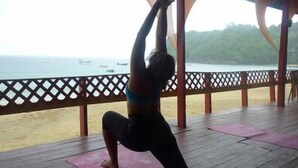 Uma pessoa fazendo uma postura de yoga em um deck de madeira, com vista para a praia e o oceano.