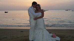 Um casal se abraçando na praia, com um mar calmo e barcos ao fundo.