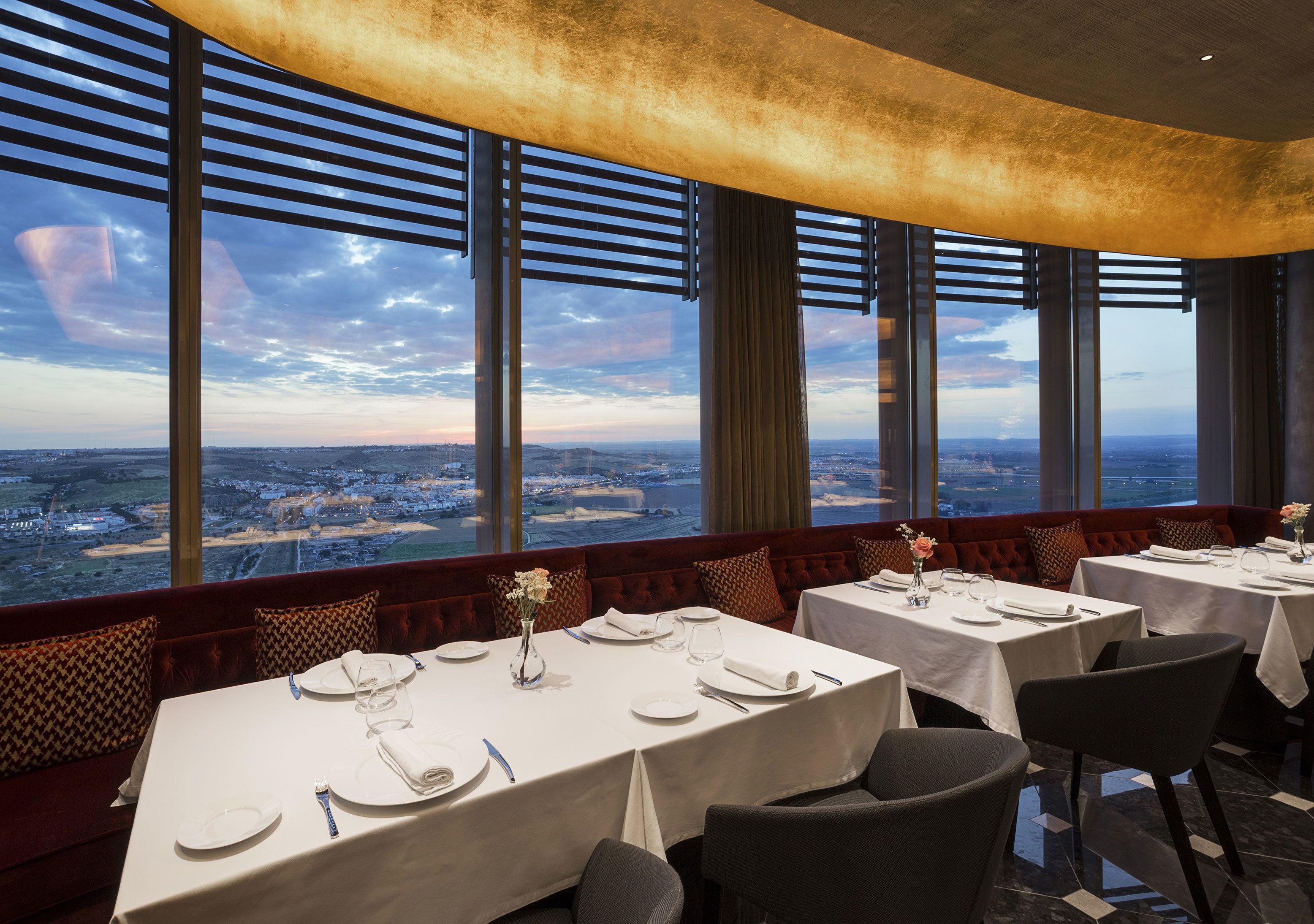 Um restaurante com mesas postas para refeições, amplas janelas com vista para a cidade e teto curvo.