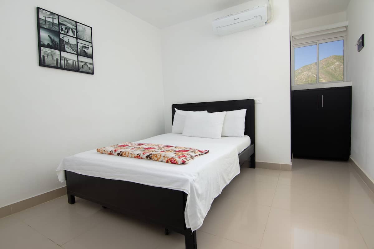 Quarto com cama, guarda-roupa preto, ar condicionado fixado na parede e janela com vista para uma montanha.