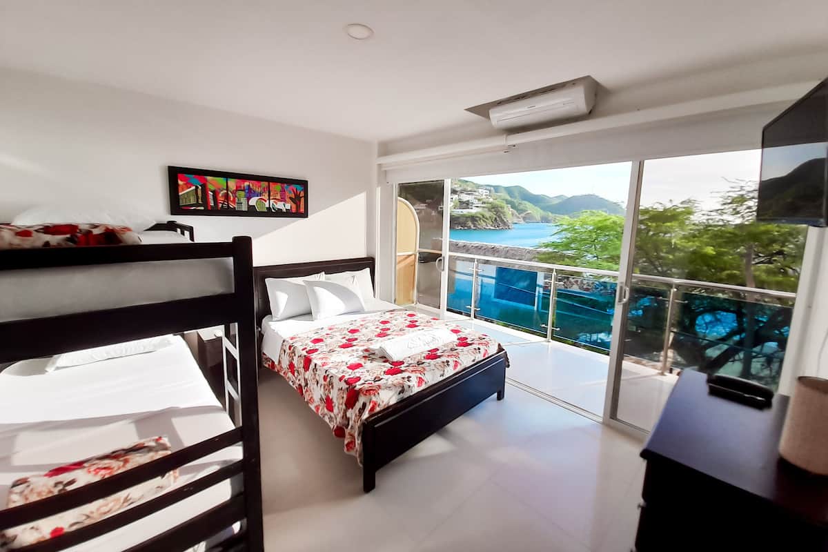 Quarto com beliche, uma cama de solteiro, uma obra de arte colorida e uma varanda com vista para o mar e colinas.