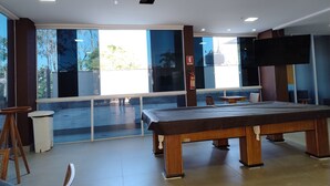 Mesa de bilhar numa sala com janelas amplas, um extintor de incêndio e uma televisão na parede.