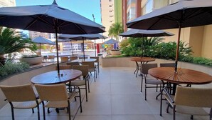 Uma área de estar ao ar livre com mesas e cadeiras redondas, protegida por guarda-sóis, cercada por vegetação e edifícios.