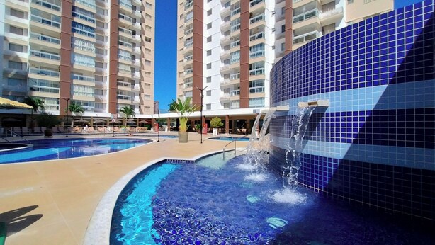 Área de piscina em resort, com um elemento aquático, cercada por edifícios altos e mobiliário de área externa.