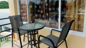 Uma varanda com mesa de vidro e quatro cadeiras, uma janela grande com cortinas e piso de azulejos.