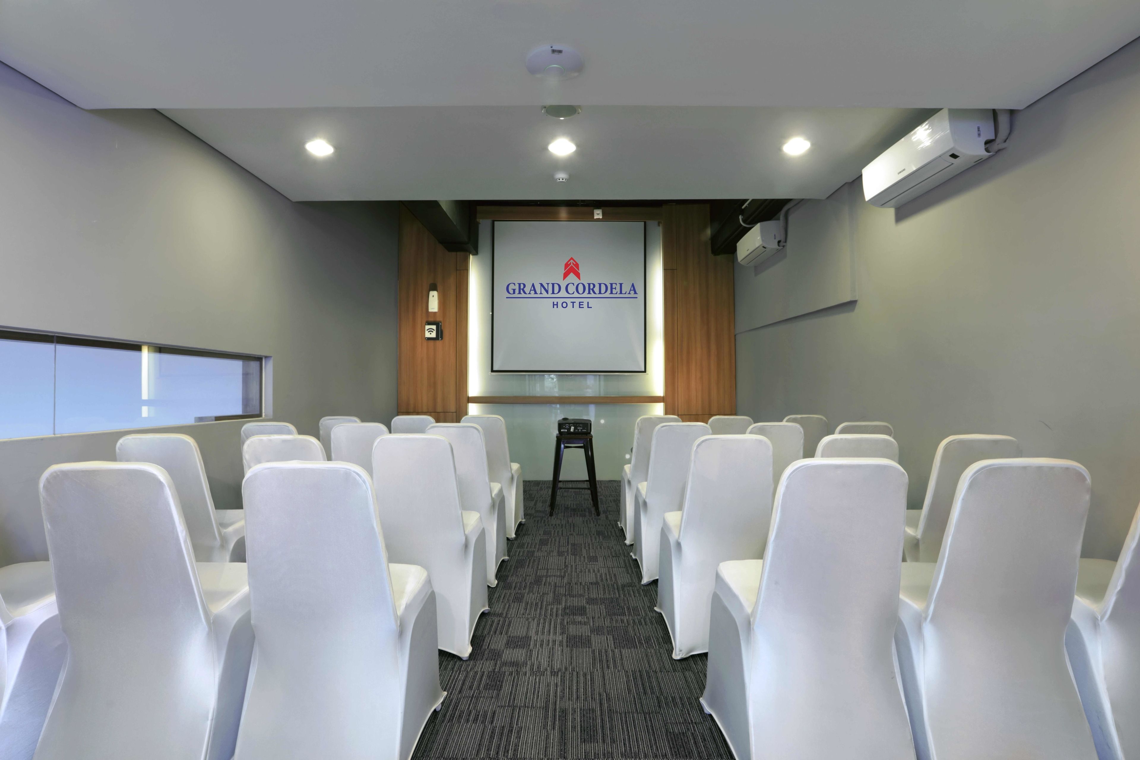 Uma sala de conferências com cadeiras brancas dispostas em fileiras, um púlpito e uma tela exibindo o logotipo do Hotel Grand Cordela.