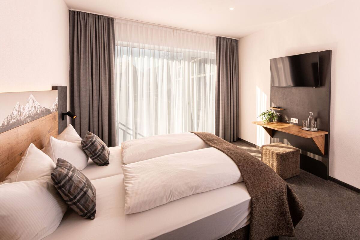 Quarto de hotel moderno com uma cama grande, televisão presa à parede, mesa de cabeceira de madeira e janela com cortinas leves.