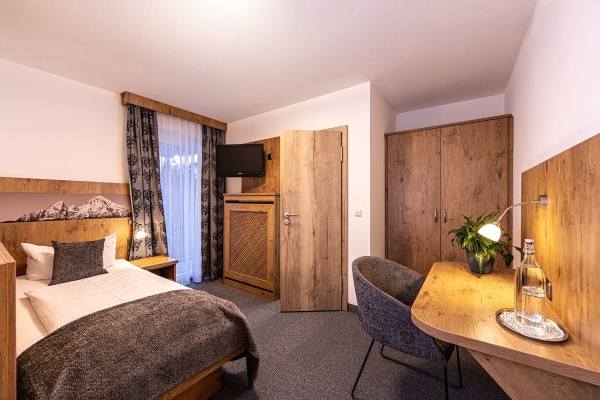 Quarto de hotel com cama, escrivaninha, cadeira, televisão e guarda-roupa.